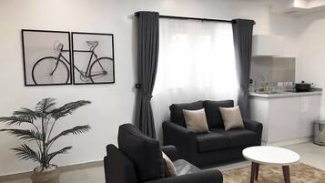 Uma sala de estar moderna com um sofá, uma mesa de centro e uma planta em vaso. Há uma área de cozinha com pia e fogão. Duas fotos emolduradas de bicicletas estão na parede.