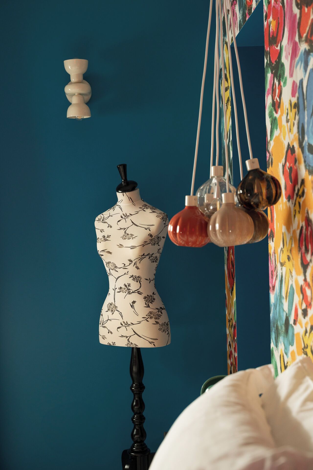 Quarto com parede azul-esverdeada, um manequim com estampas florais e uma cama com roupa de cama branca.