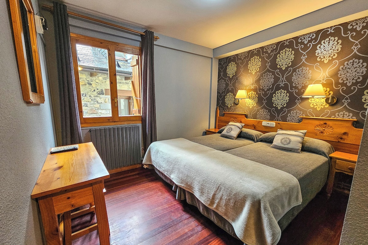 Quarto com cama de madeira, duas mesas de cabeceira, um criado-mudo, janela com cortinas e parede estampada.
