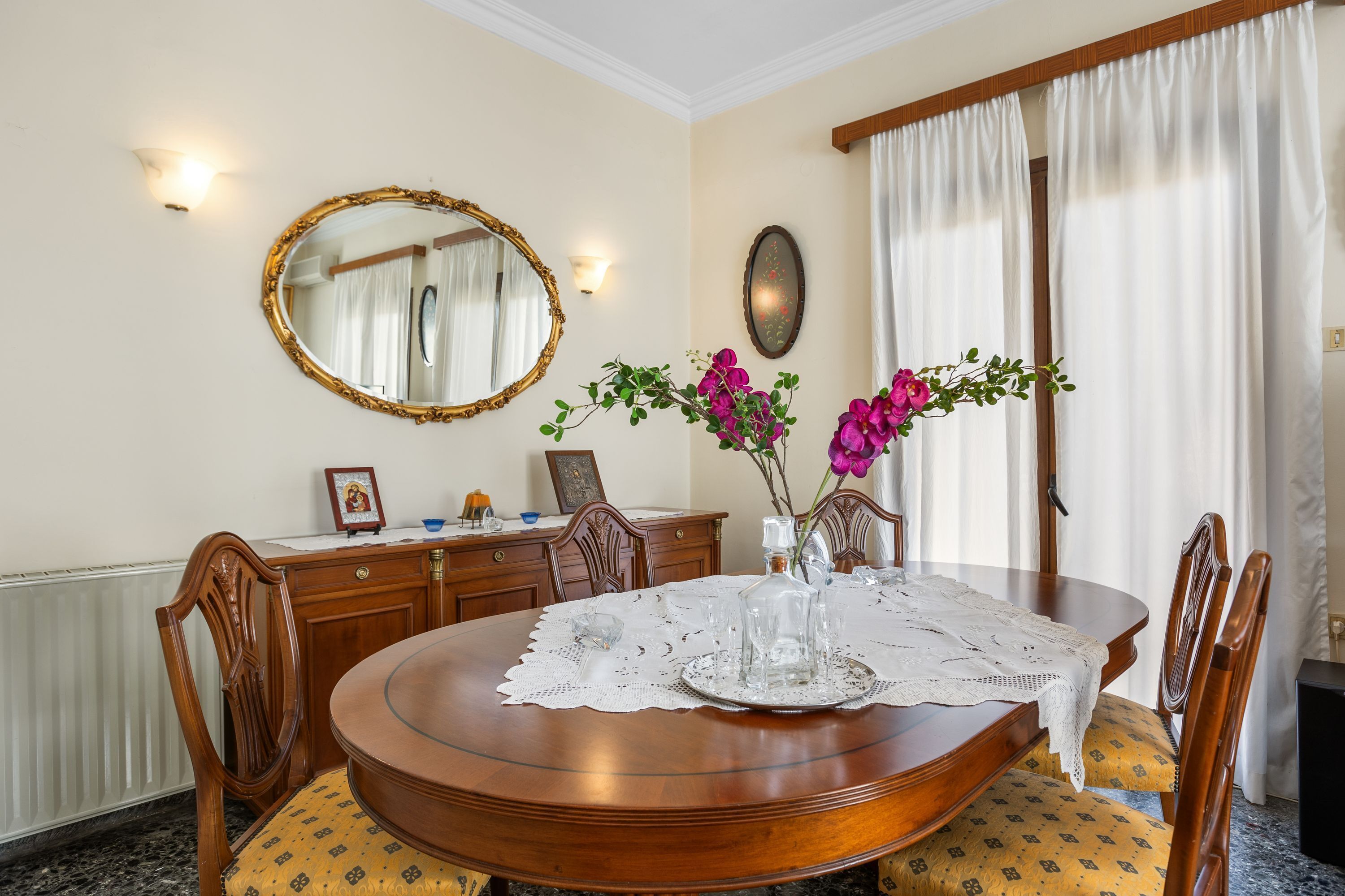 Uma sala de jantar com uma mesa redonda de madeira, toalha rendada e cadeiras com almofadas estampadas.
