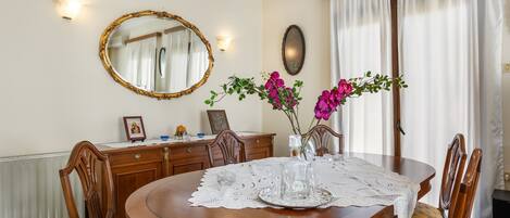 Uma sala de jantar com uma mesa redonda de madeira, toalha rendada e cadeiras com almofadas estampadas.