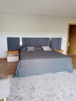 Quarto moderno com uma cama grande, mesa de cabeceira de madeira e cabeceira cinza.