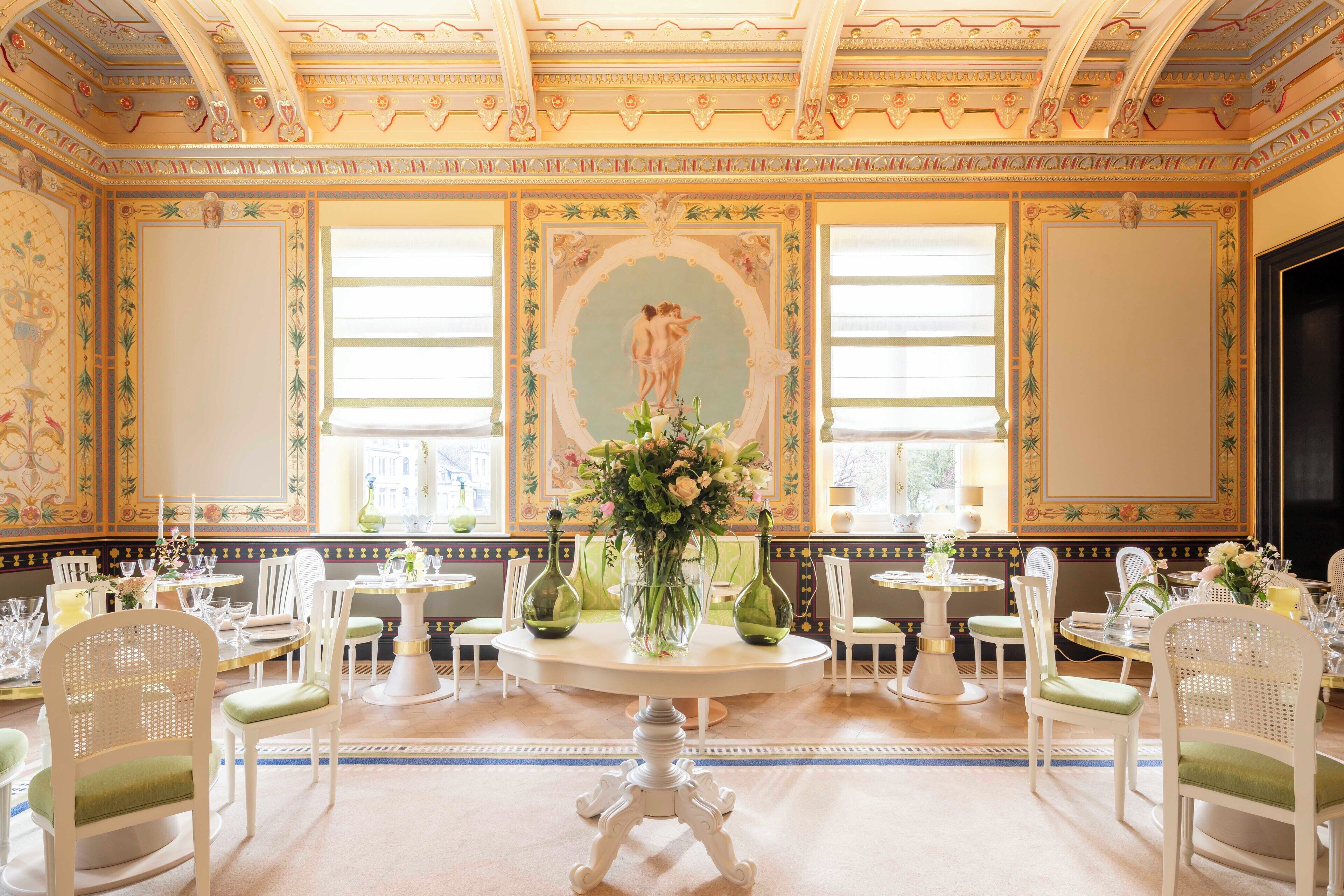 Uma sala de jantar formal, com decoração ornamentada no teto e nas paredes, um arranjo floral central e várias mesas de jantar com cadeiras.