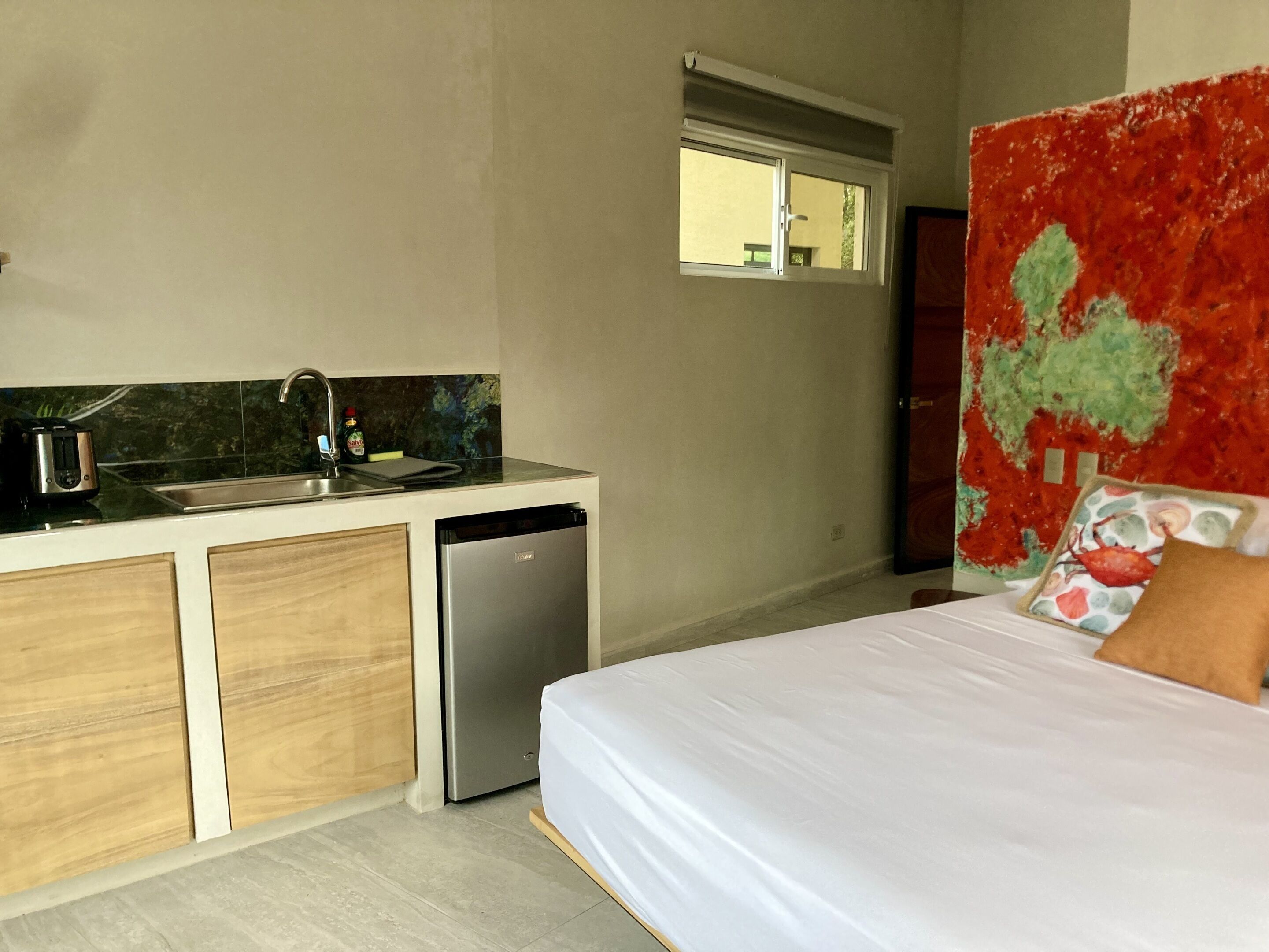 Quarto compacto com cama, pequena cozinha equipada com pia e máquina de lavar louça, e uma janela.