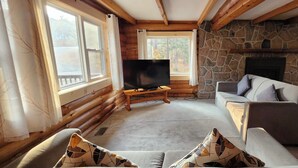 Interior de uma cabana de tronco com lareira de pedra, televisão sobre um suporte de madeira e um sofá cinza com almofadas estampadas.