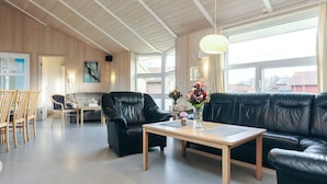 Interior moderno com teto de madeira, sofá de couro preto, mesa de madeira com um vaso de flores e uma área de refeições com cadeiras e uma mesa pequena.