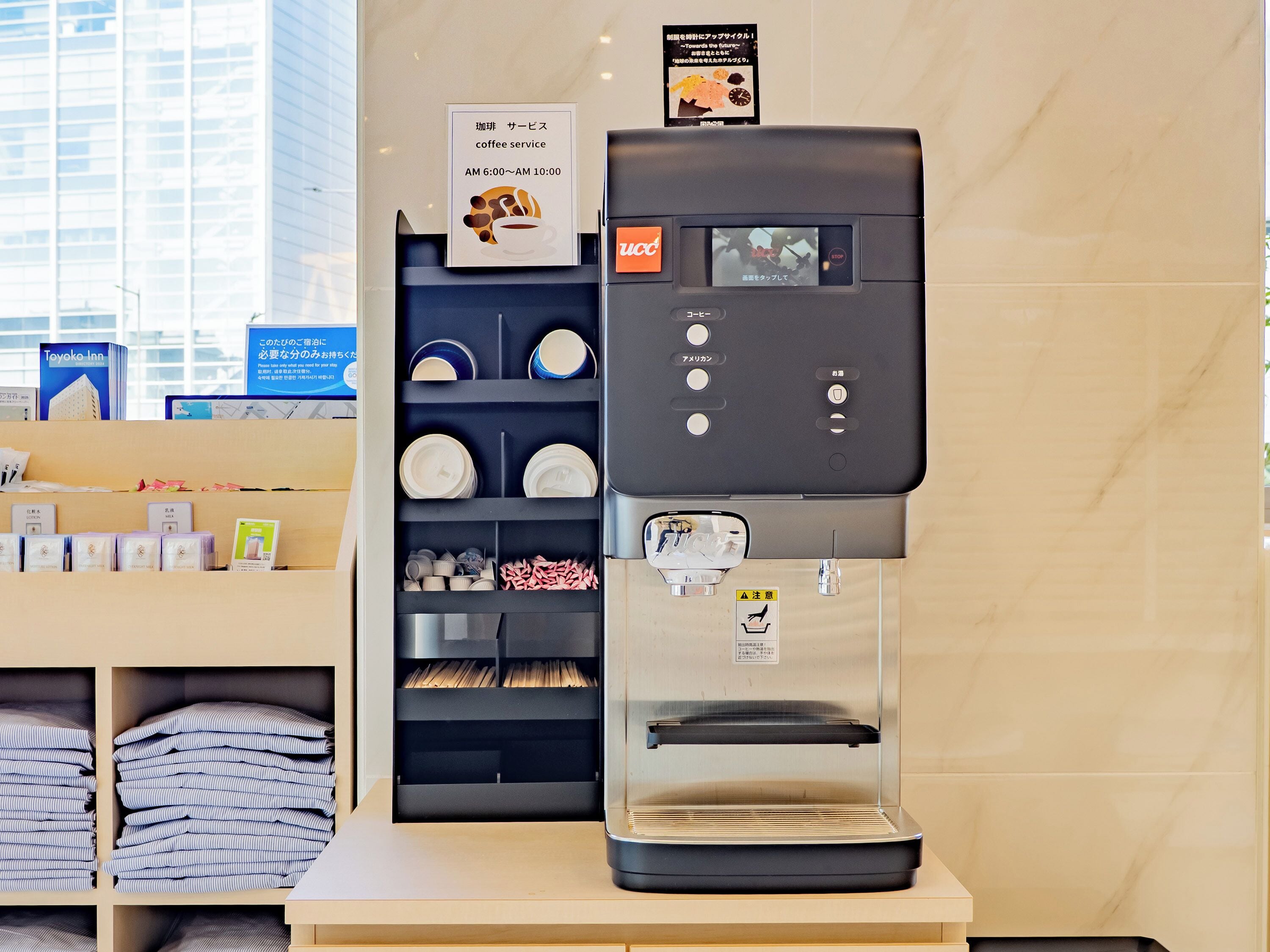 Máquina de café com visor digital e botões, placa com horário de funcionamento do serviço de café, e prateleiras com xícaras e outros objetos.