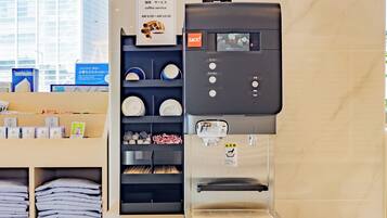 Máquina de café com visor digital e botões, placa com horário de funcionamento do serviço de café, e prateleiras com xícaras e outros objetos.