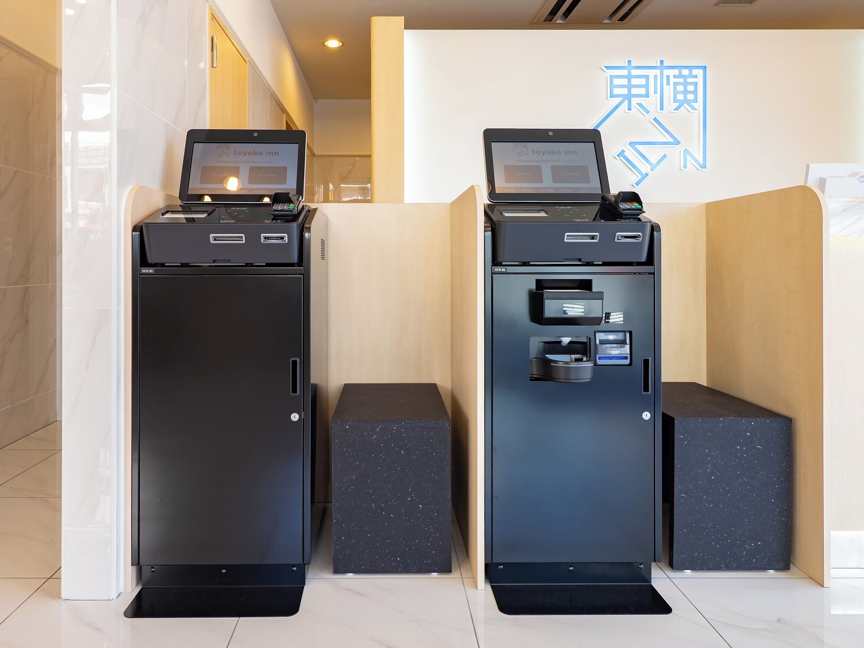 Dois totens de autoatendimento com terminais de pagamento e visor digital, provavelmente para registo ou serviço de quartos, num hall moderno de hotel.