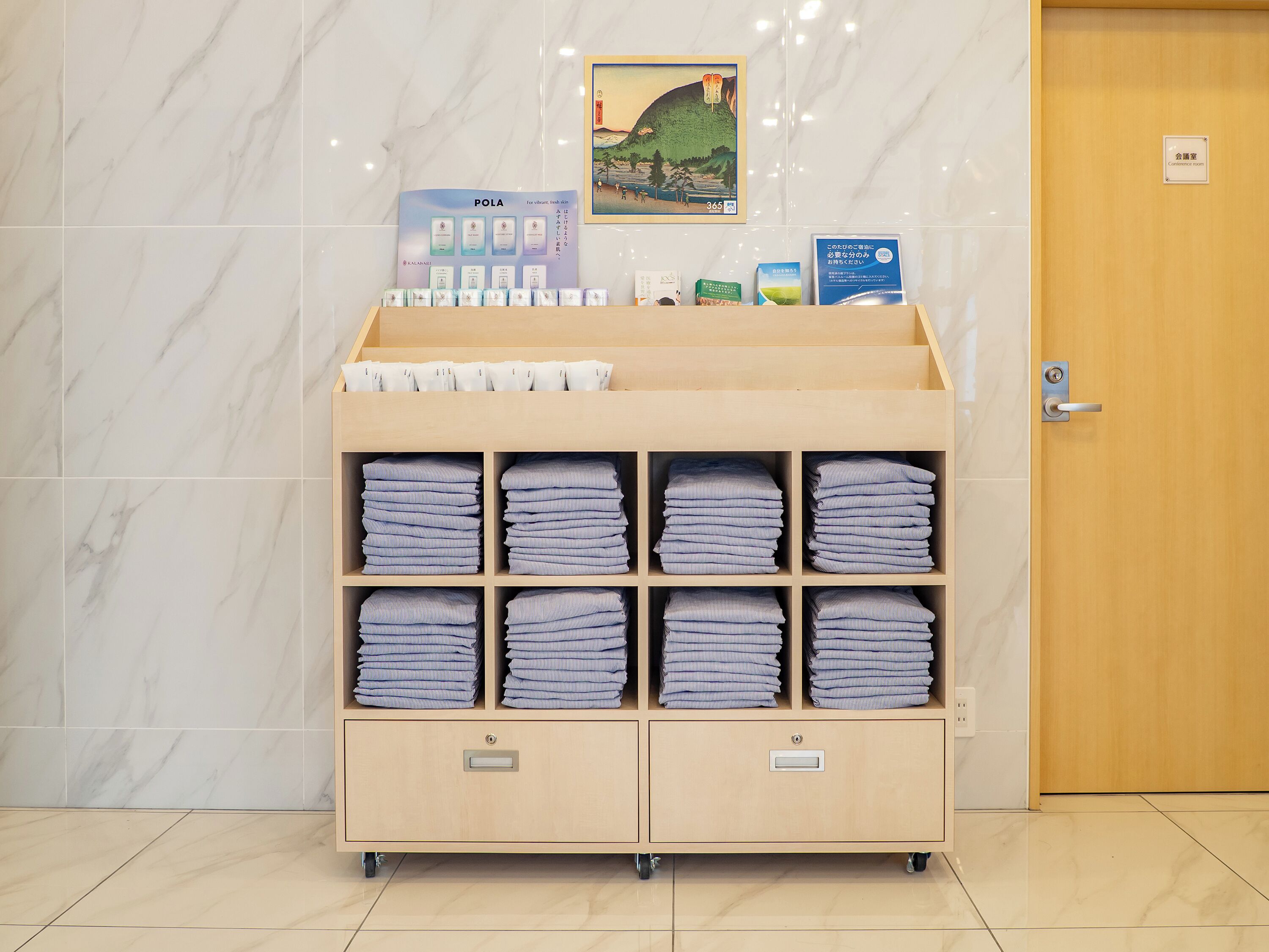 Uma área de recepção de hotel com parede de mármore, vitrine de madeira com toalhas dobradas, quadro emoldurado e placa indicativa da sala de conferências.