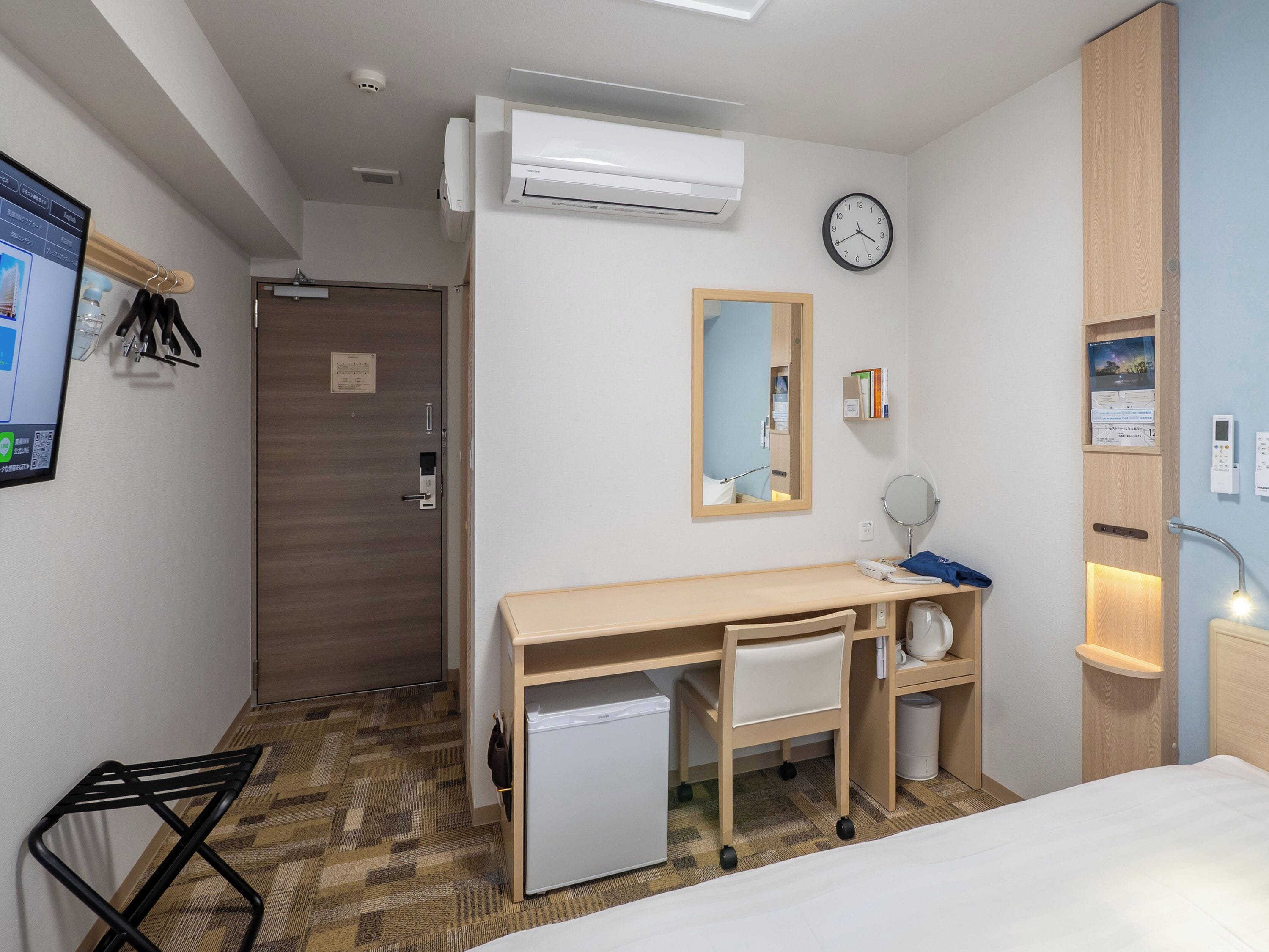 Quarto de hotel com cama, escrivaninha com cadeira, pequeno frigorífico, ar condicionado, relógio, espelho e prateleira com luminária.