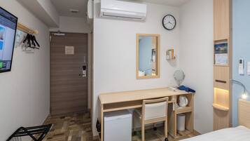 Quarto de hotel com cama, escrivaninha com cadeira, pequeno frigorífico, ar condicionado, relógio, espelho e prateleira com luminária.
