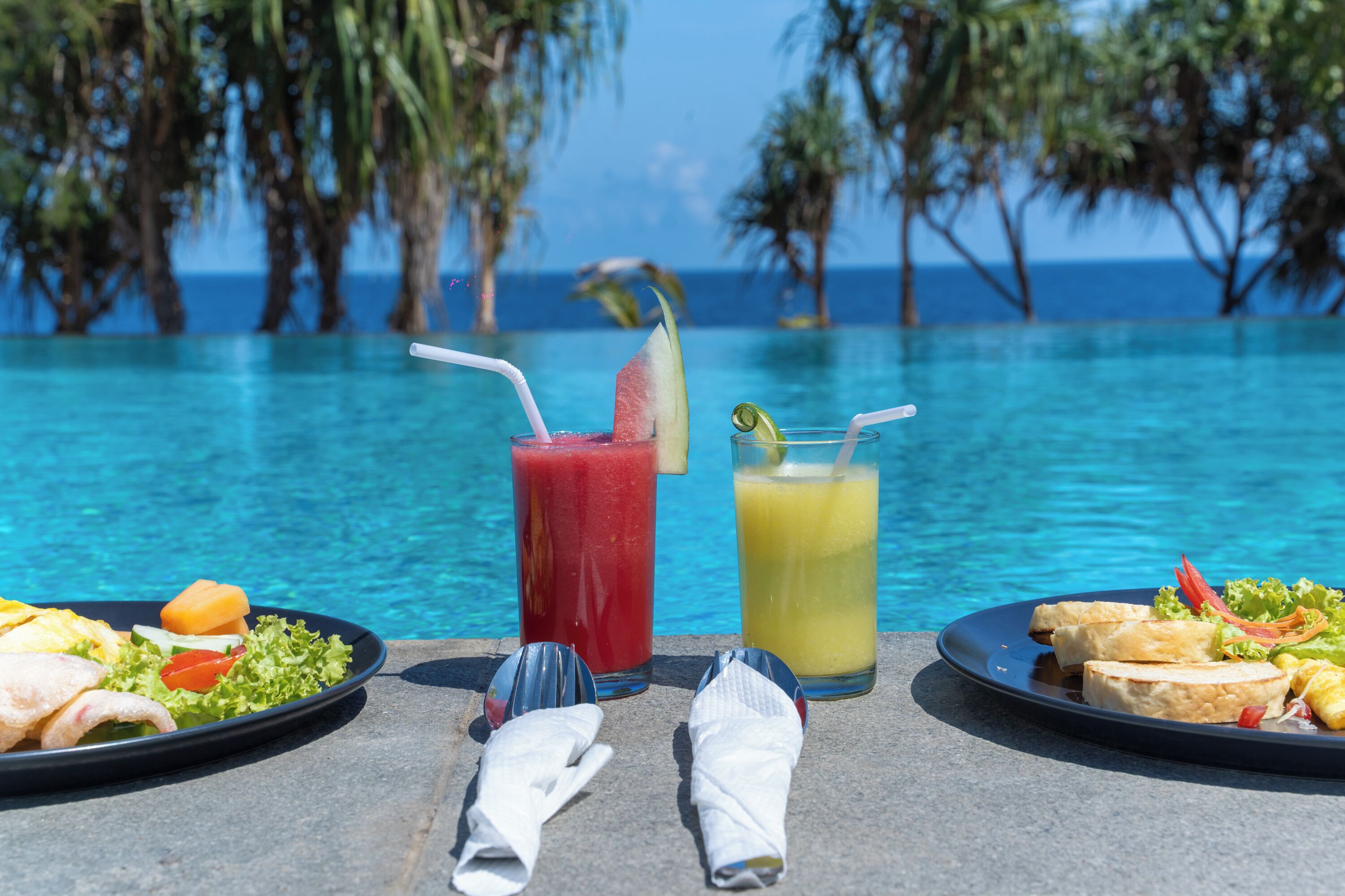 Mesa de jantar com pratos de comida e bebidas à beira da piscina, com vista para o oceano.