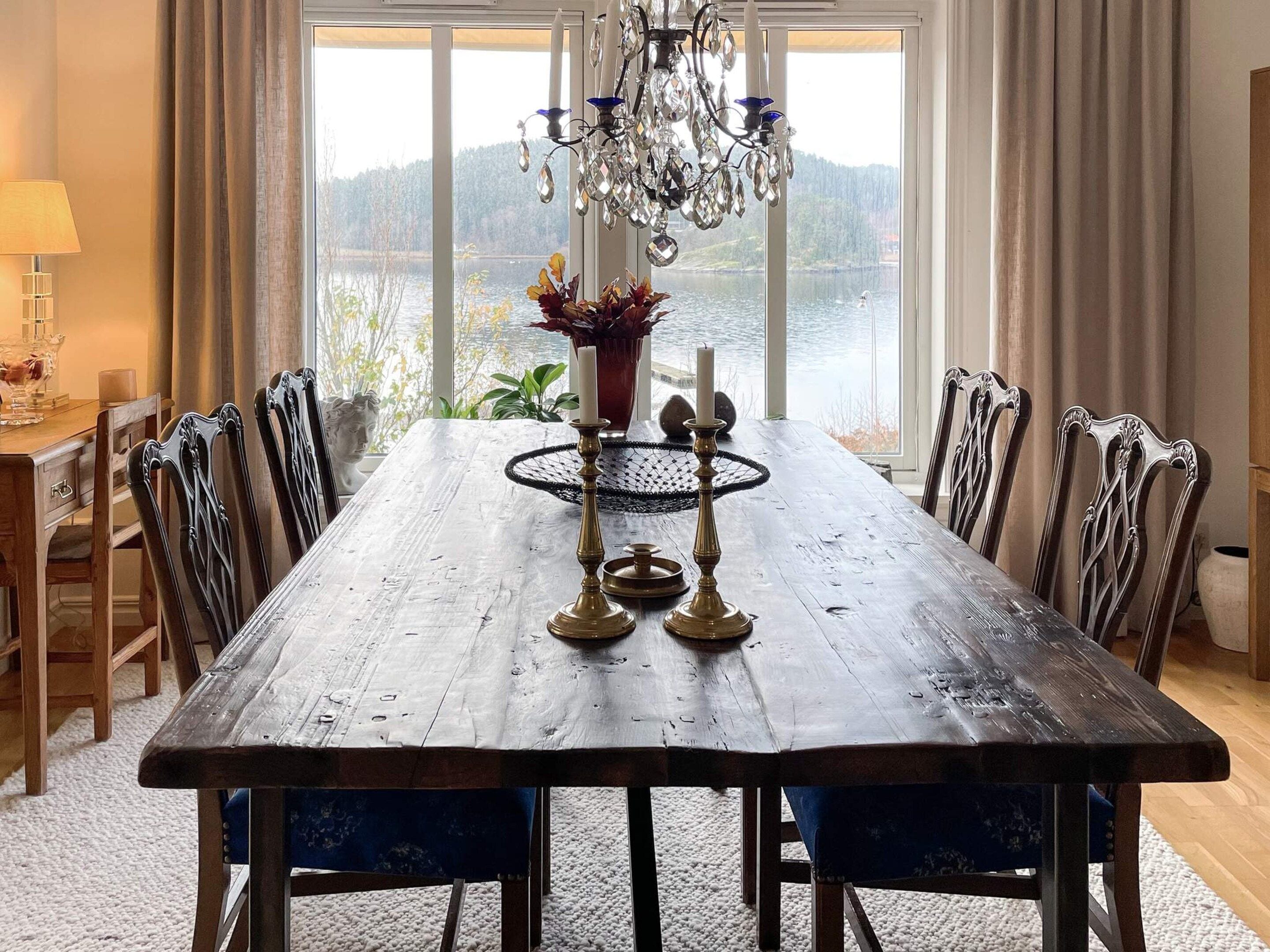 Uma sala de jantar com uma grande mesa de madeira, cadeiras elaboradas, um candelabro e vista para um lago através de amplas janelas.