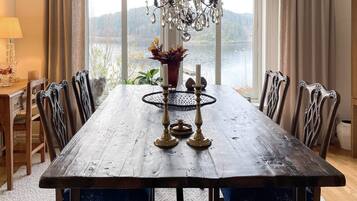 Uma sala de jantar com uma grande mesa de madeira, cadeiras elaboradas, um candelabro e vista para um lago através de amplas janelas.