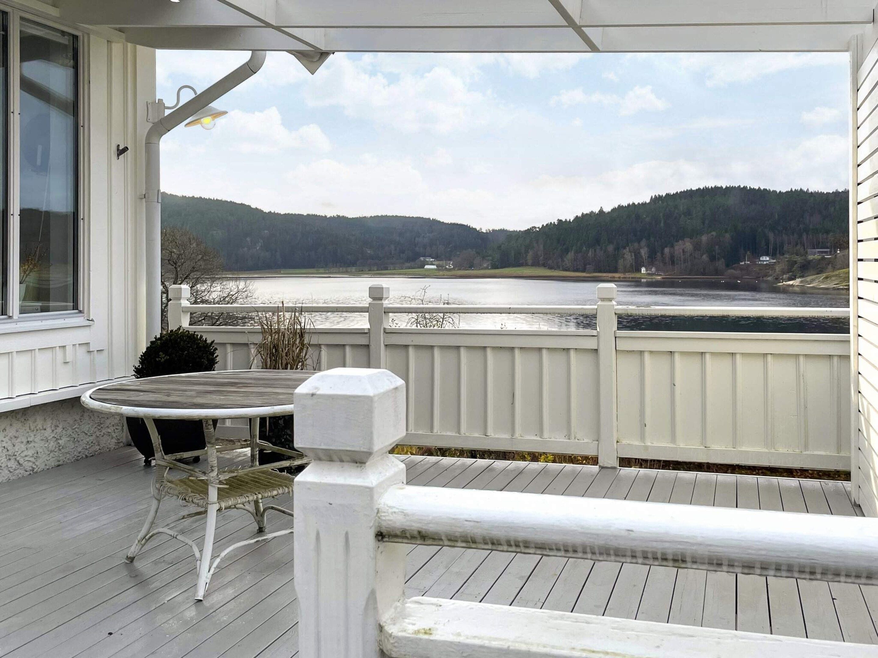 Um deck coberto ao ar livre com mesa e cadeiras, com vista para um lago e colinas arborizadas.