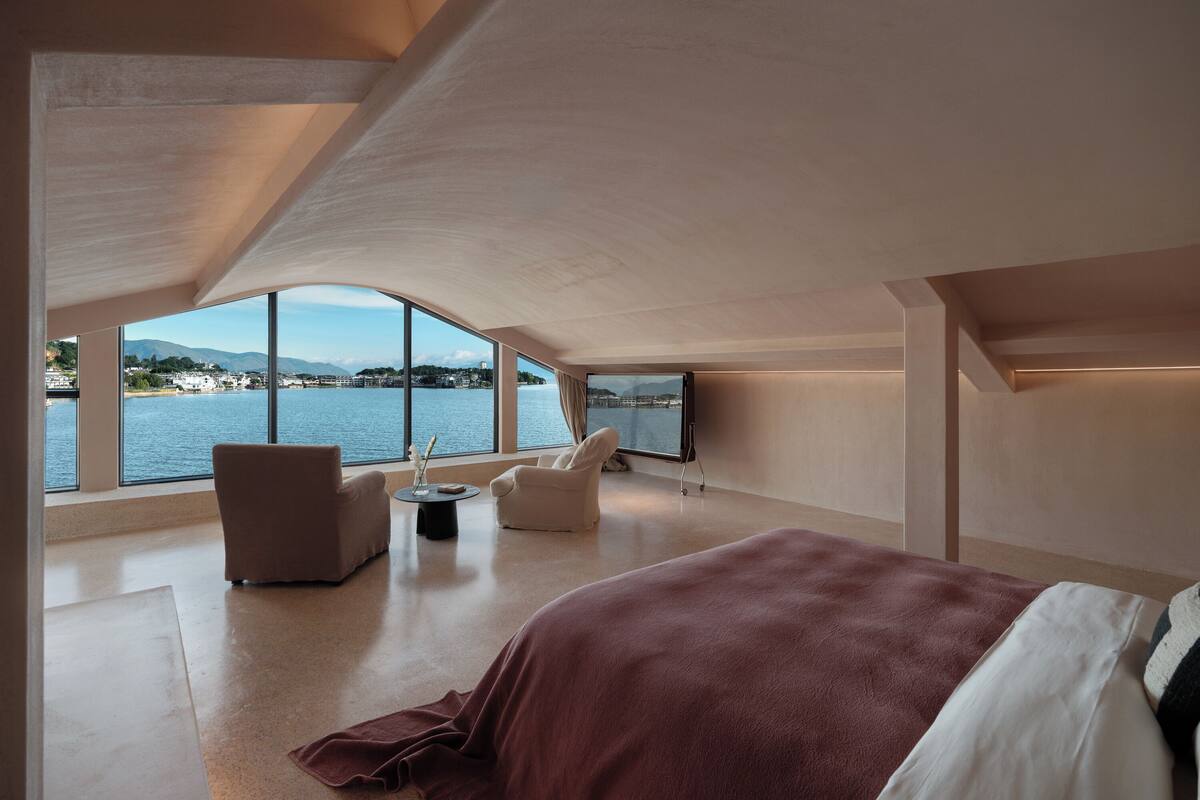 Quarto moderno com uma cama grande, uma escrivaninha e uma cadeira, com vista para um corpo d'água e o horizonte da cidade.