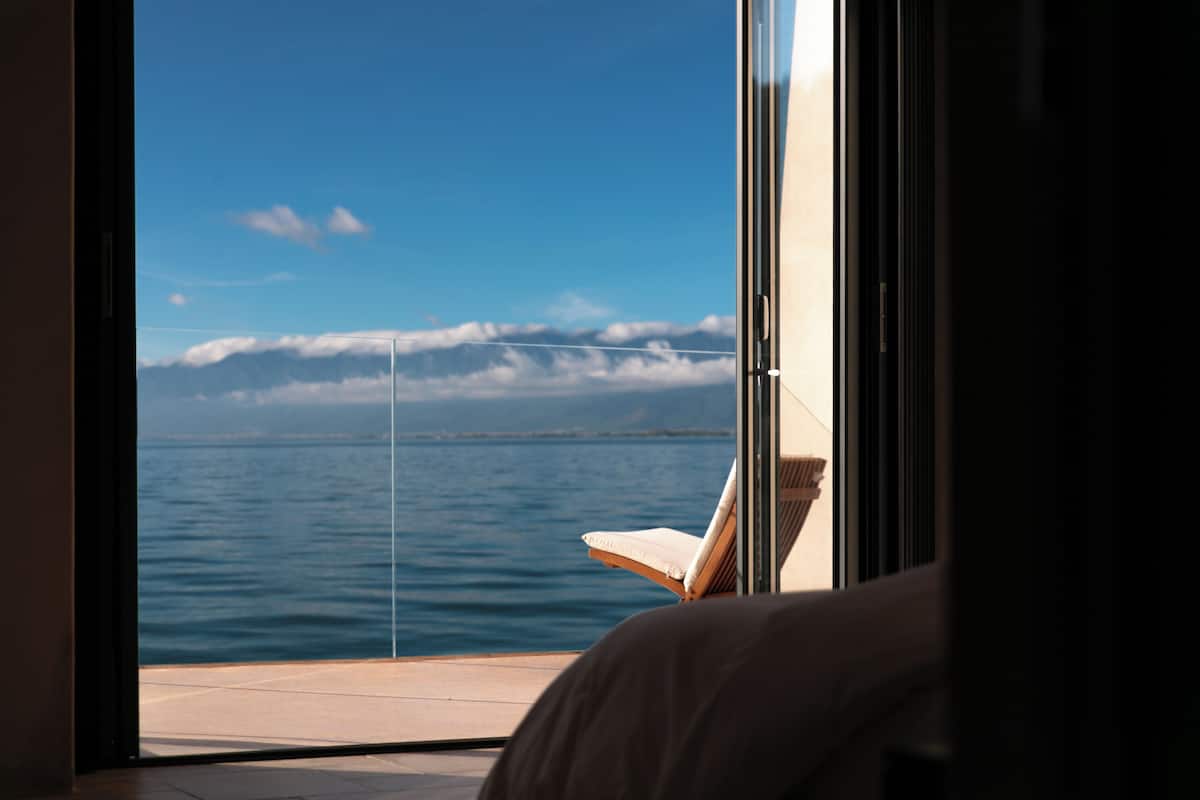 Quarto com uma grande janela com vista para um corpo d'água e montanhas ao longe.