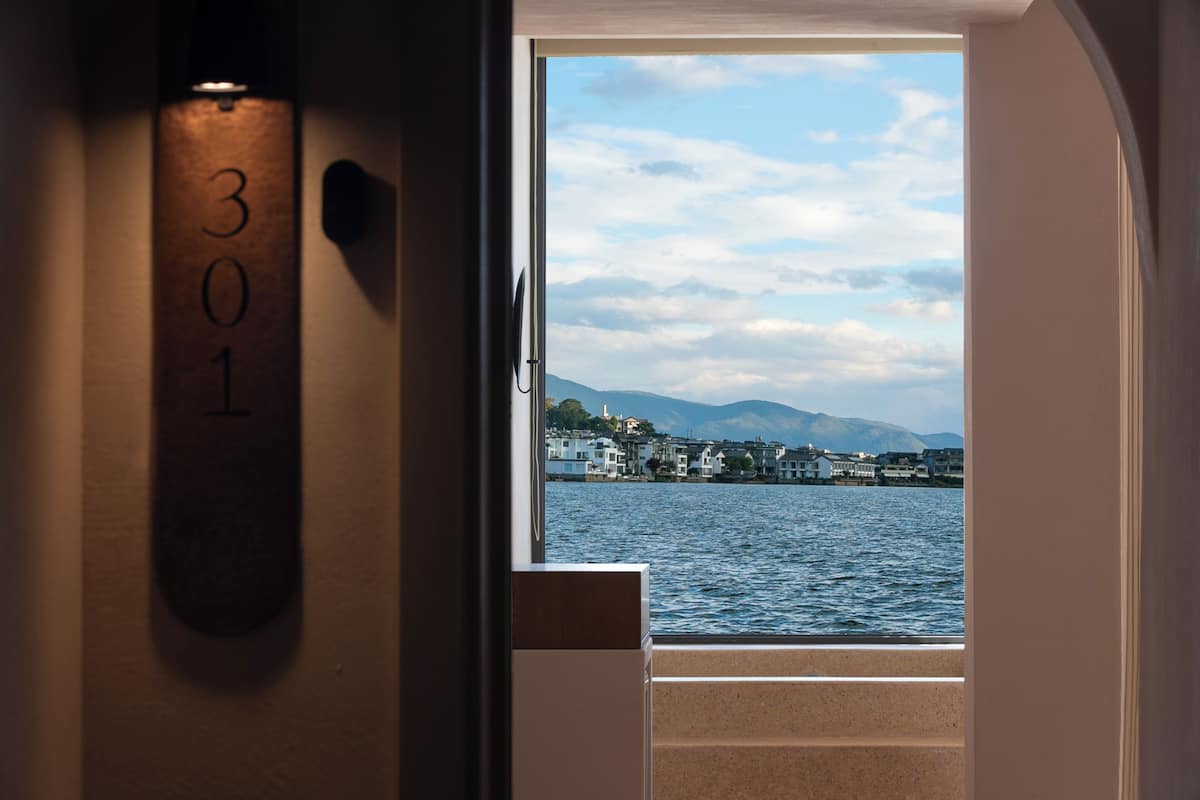 Uma porta com uma placa de identificação com o número "301", dando acesso a uma vista de um corpo d'água, com edifícios e montanhas ao longe.