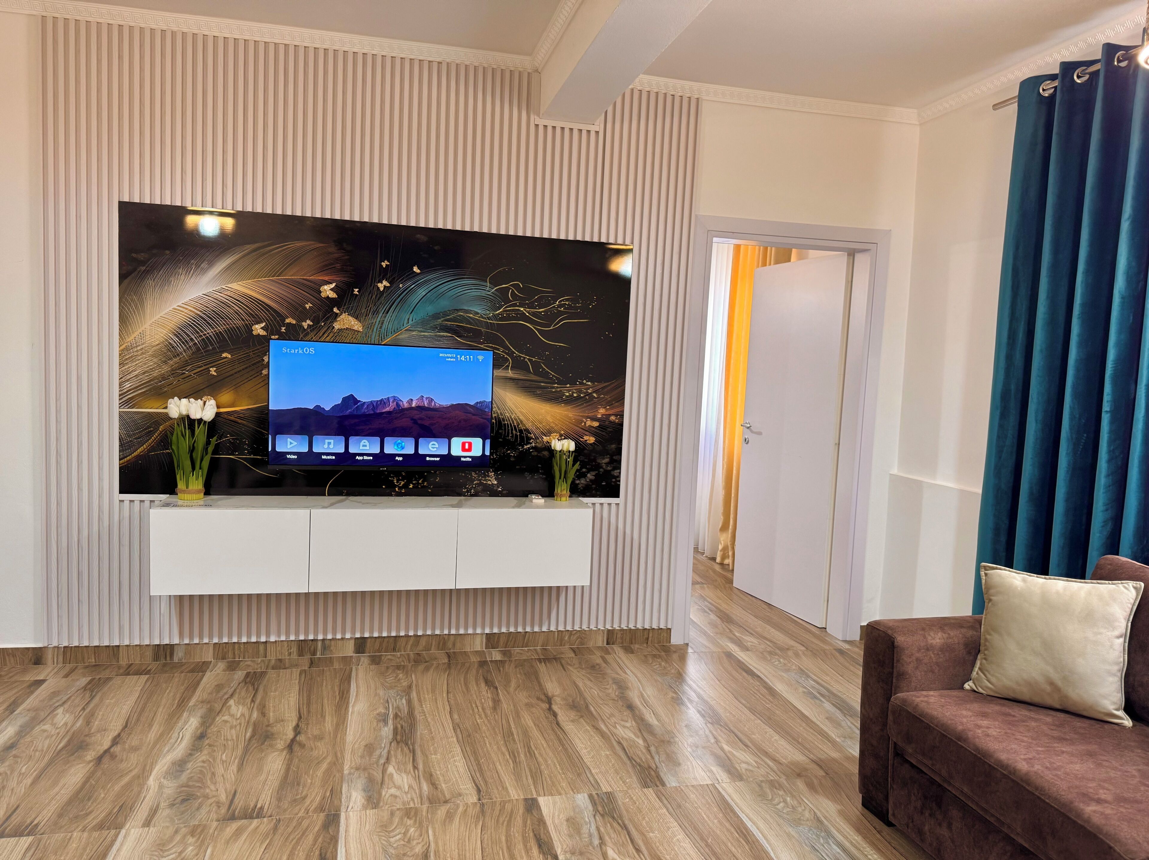 Uma sala de estar com TV na parede, piso de madeira, sofá marrom e uma porta com cortina azul.