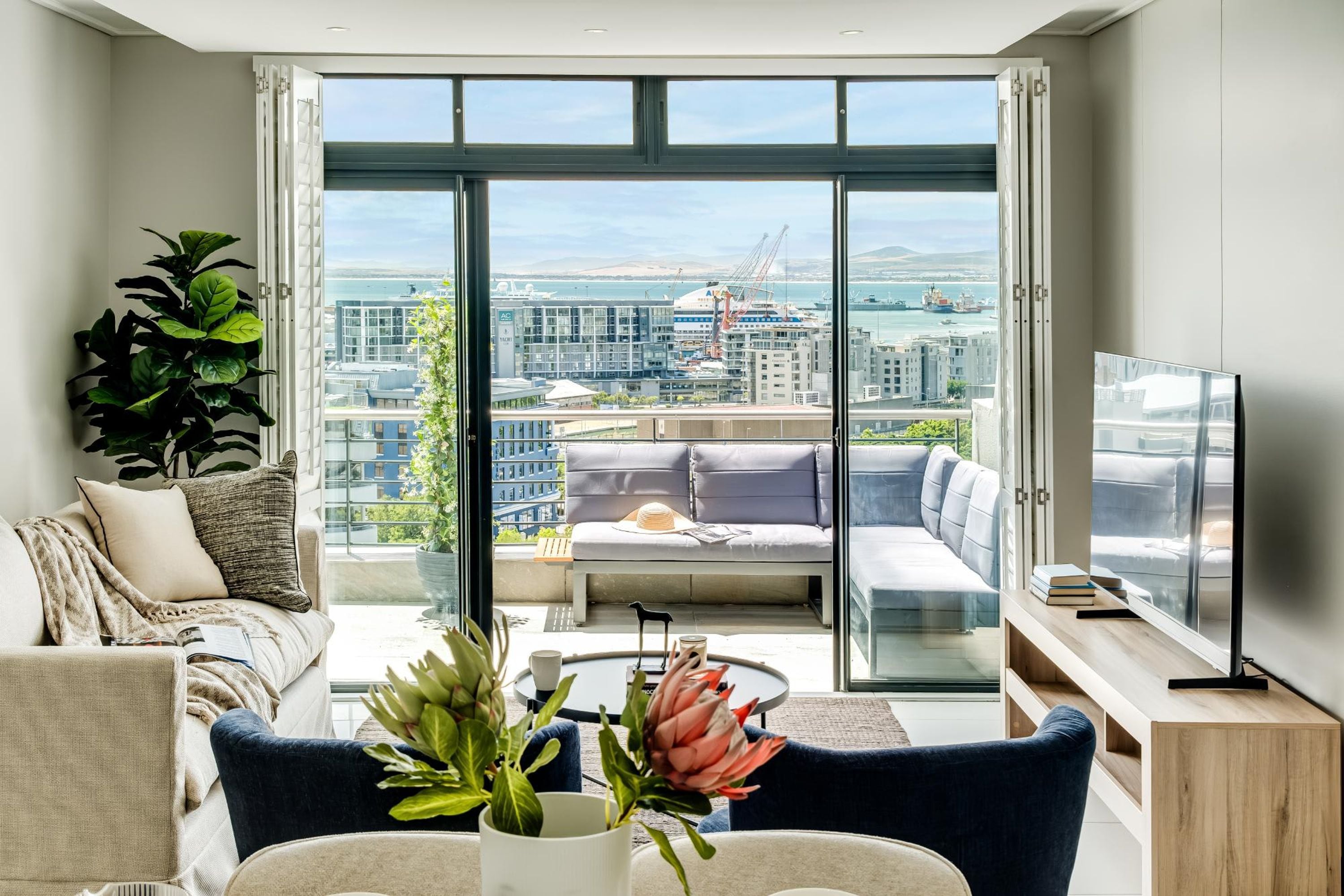 Sala de estar moderna com sofá, mesa de centro e televisão. Uma janela ampla oferece vista para a paisagem urbana e para a água.