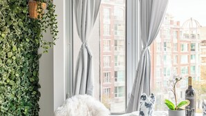 Quarto moderno com uma mesa redonda, uma poltrona macia, uma parede verde e vista para prédios através de janelas grandes com cortinas cinzentas.