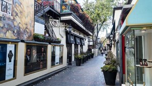 Rua estreita com lojas e cafés charmosos, com mesas ao ar livre e plantas em vasos.