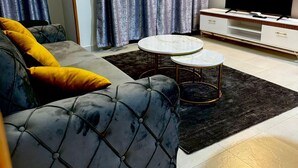 Uma sala de estar moderna com um sofá cinza, uma mesa de centro redonda pequena, uma televisão de tela plana em um suporte branco e uma cortina estampada.