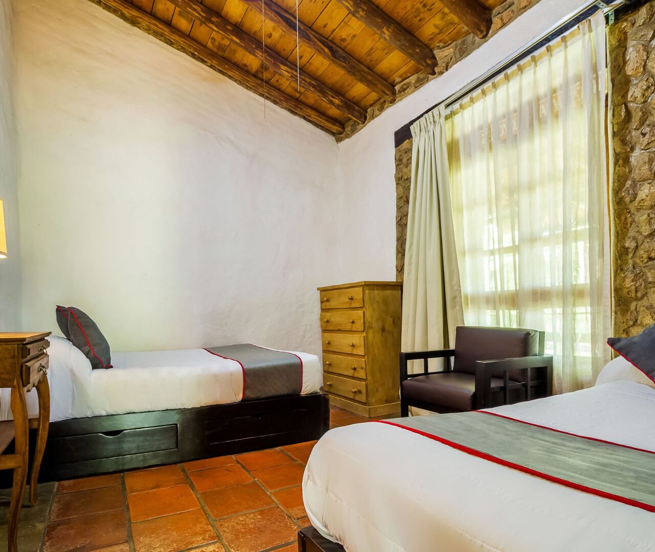 Quarto com duas camas, cômoda de madeira, cadeira e janela com cortinas.