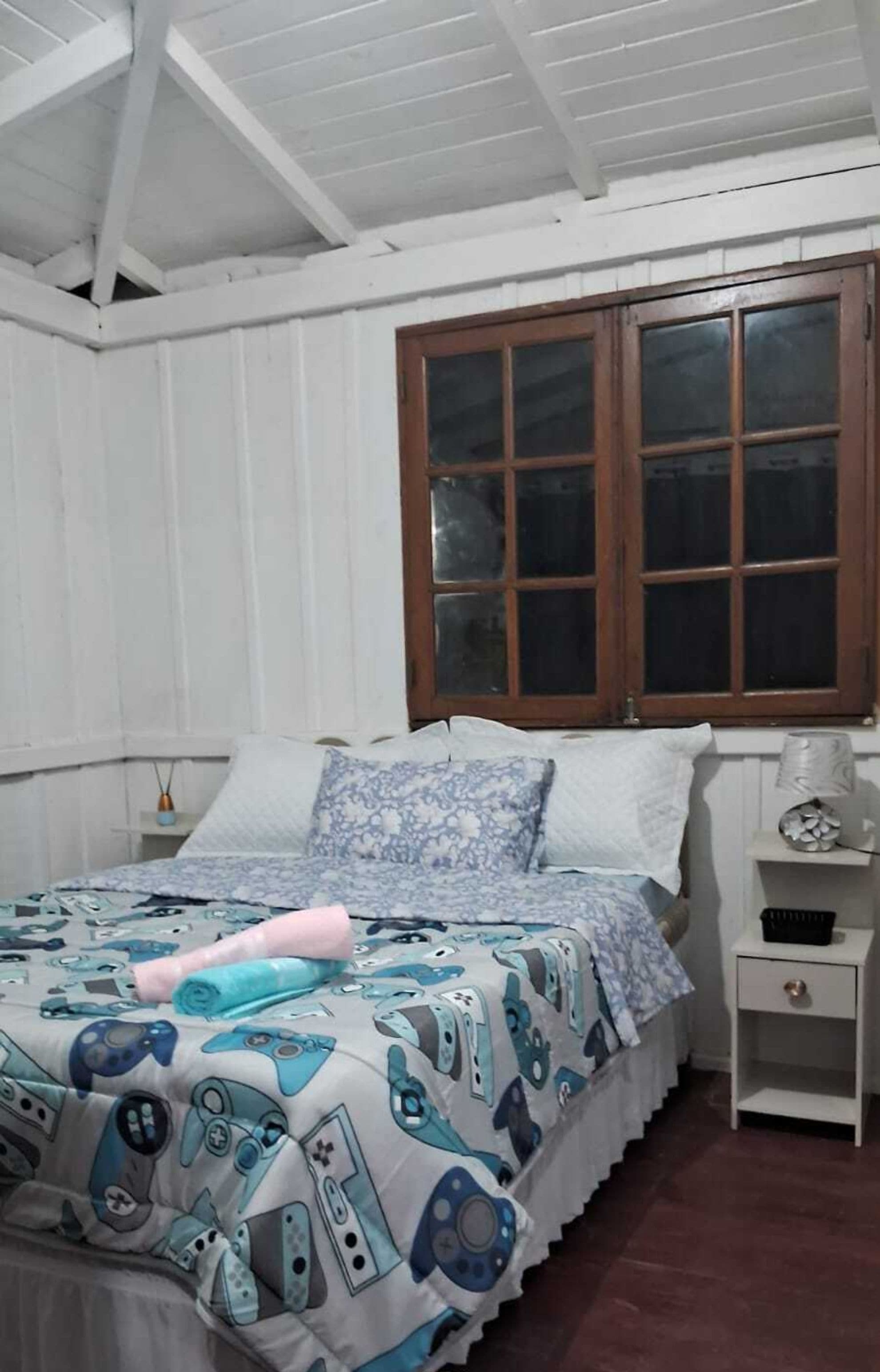 Quarto com cama, mesa de cabeceira, janela e teto com vigas aparentes.