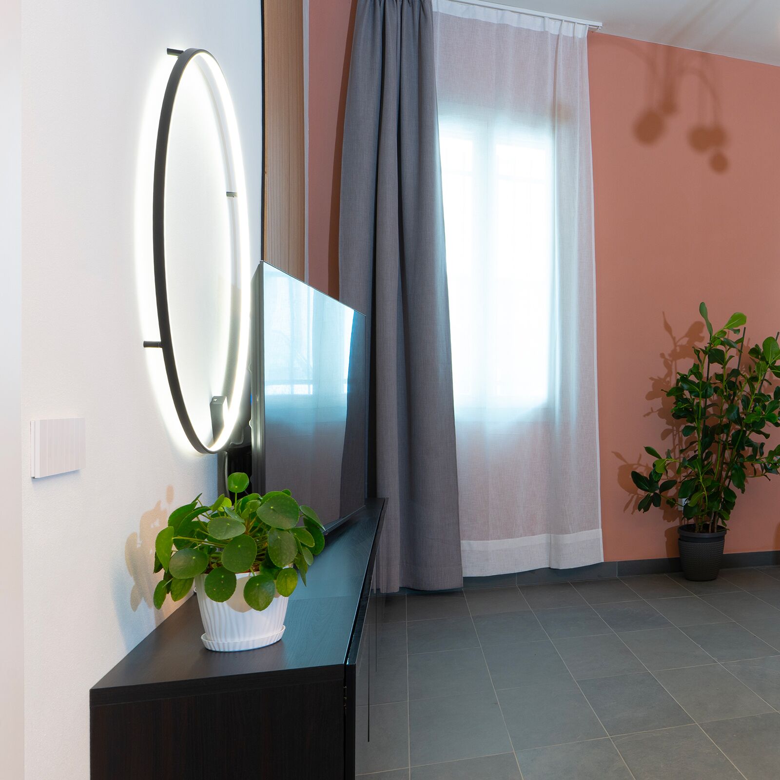 Quarto moderno com televisão de tela plana presa a um armário escuro, uma planta em vaso sobre o balcão e uma grande janela com cortinas.