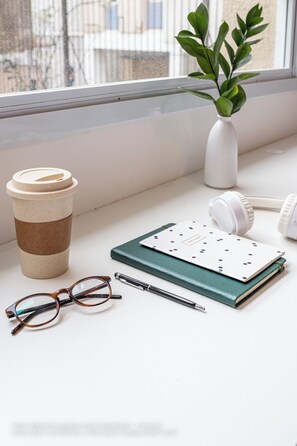 Um peitoril de janela com uma xícara de café, um par de óculos, um caderno, uma caneta, fones de ouvido e um vaso com planta.