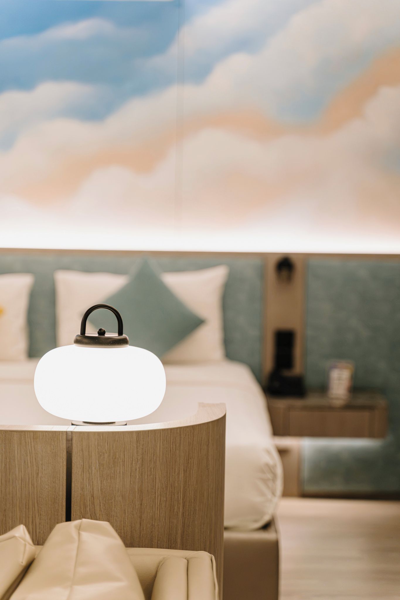 Uma cama com cabeceira, uma mesa de cabeceira com abajur, e uma parede com pintura do céu e nuvens.