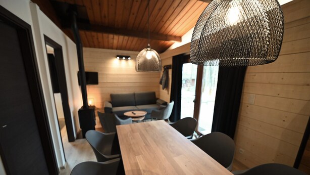 Uma área de refeições moderna com mesa e cadeiras de madeira, luminária pendente e um sofá ao fundo.