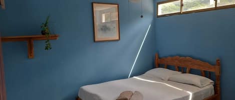 Um quarto com uma cama de solteiro, um ventilador de teto, uma janela com vista e um quadro na parede.