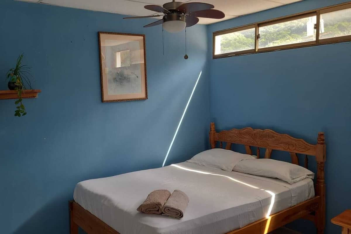 Um quarto com uma cama de solteiro, um ventilador de teto, uma janela com vista e um quadro na parede.