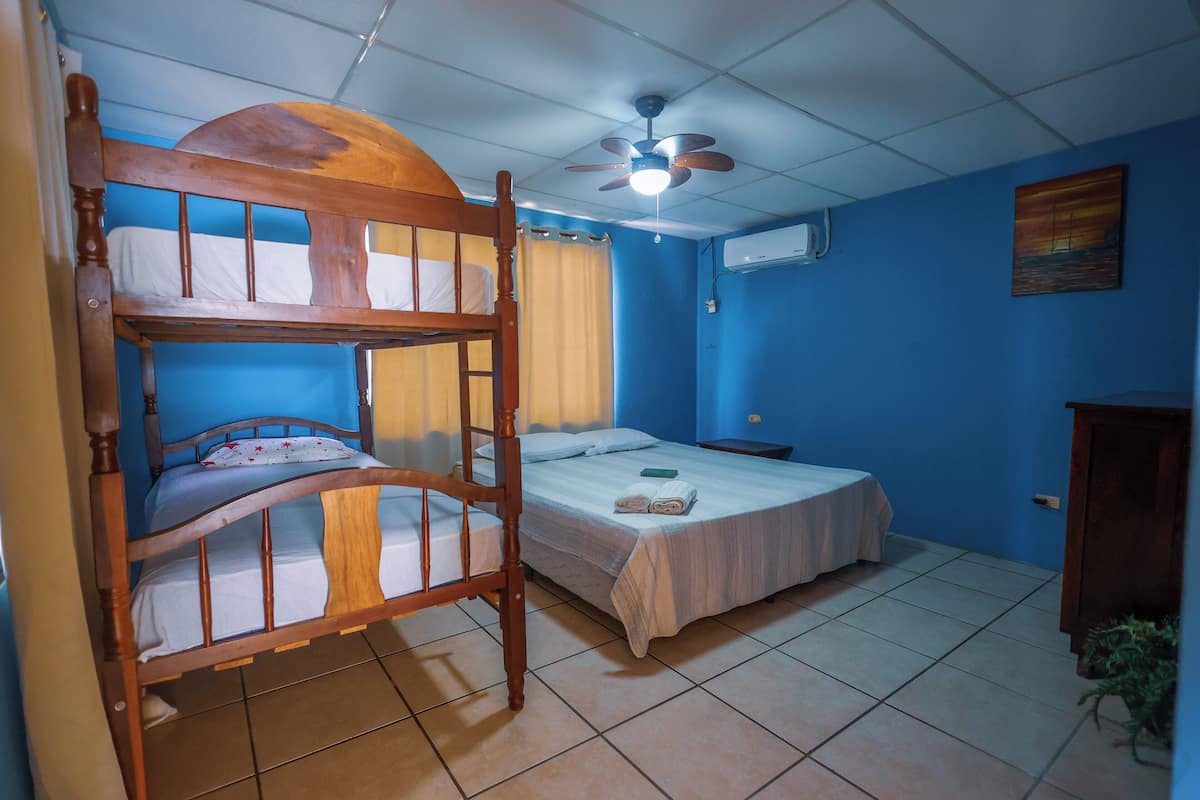 Um quarto com cama de solteiro, uma cama individual, ventilador no teto e janela com cortinas.