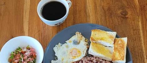 Uma refeição com arroz frito, duas fatias de torrada, um ovo frito e uma pequena tigela de legumes picados, sobre um prato cinzento, com uma xícara de café preto em uma mesa de madeira.