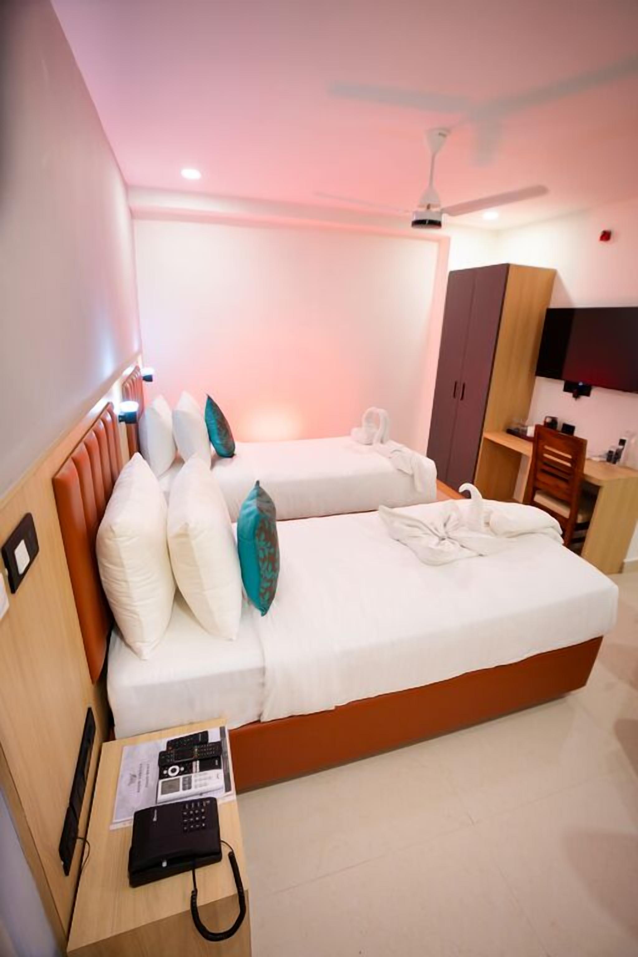 Quarto de hotel com duas camas, uma escrivaninha, uma cadeira, uma televisão e ventoinha de teto.