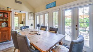 Uma sala de jantar com uma mesa de madeira, seis cadeiras estofadas e vista para um pátio externo através de grandes portas de vidro.