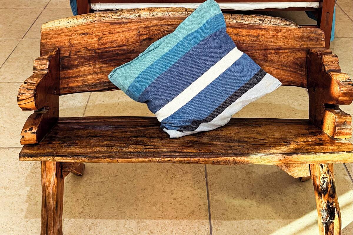 Uma cama com travesseiro listrado, um banco de madeira, uma mesinha de cabeceira com abajur e uma prateleira com objeto decorativo.