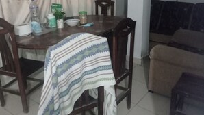 Uma área de jantar com uma mesa posta para quatro pessoas, um dispensador de água, uma planta em vaso e uma tigela. Há um sofá e uma cadeira ao fundo.