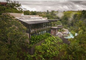Um edifício moderno com fachada de vidro, rodeado por vegetação exuberante e área de piscina.