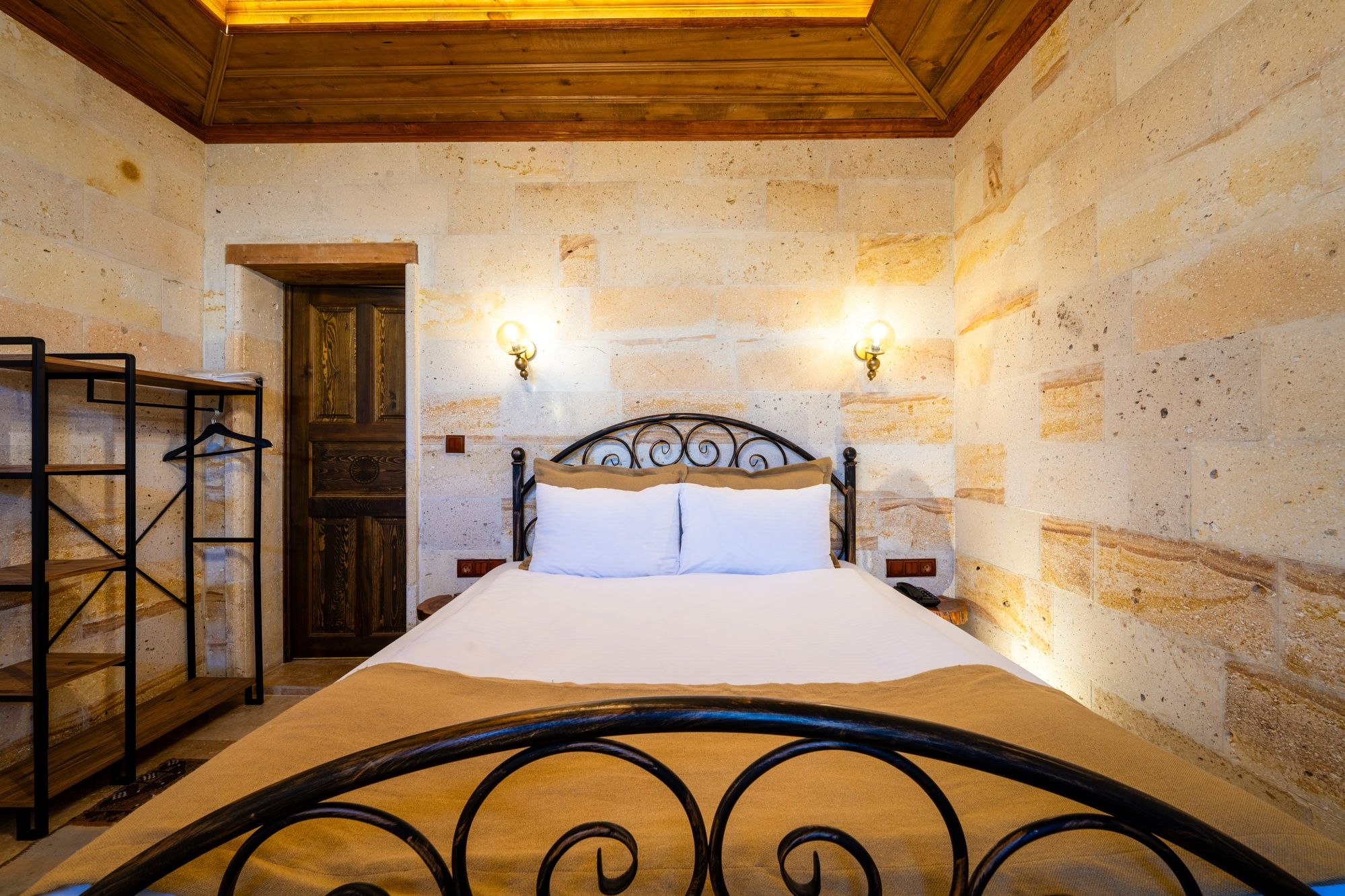 Quarto com cama, cabeceira de madeira, paredes de pedra e teto de madeira.