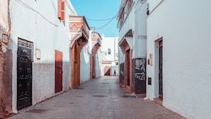 Uma rua estreita de paralelepípedos ladeada por edifícios brancos com portas e janelas vermelhas, conduzindo a um arco distante.