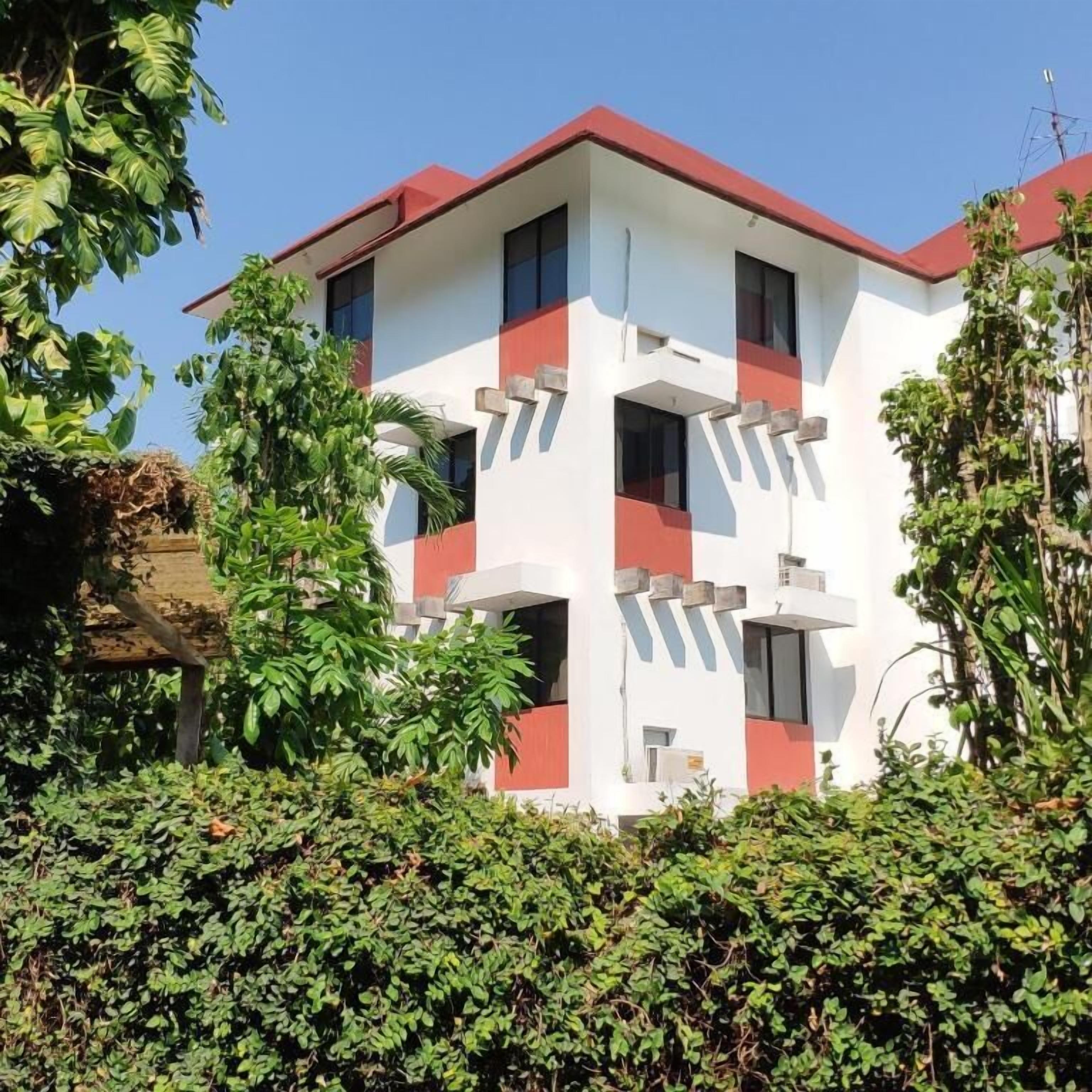 Um edifício de vários andares com telhado vermelho e paredes brancas, rodeado por vegetação exuberante.