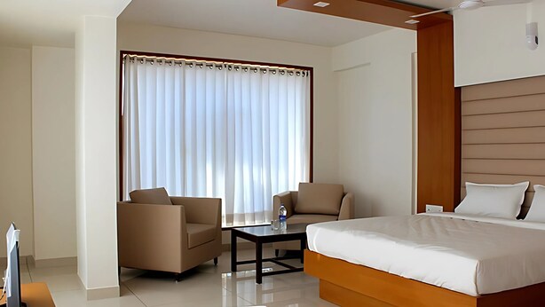 Quarto de hotel moderno com uma cama grande, duas poltronas, uma mesa pequena e uma janela ampla com cortinas.