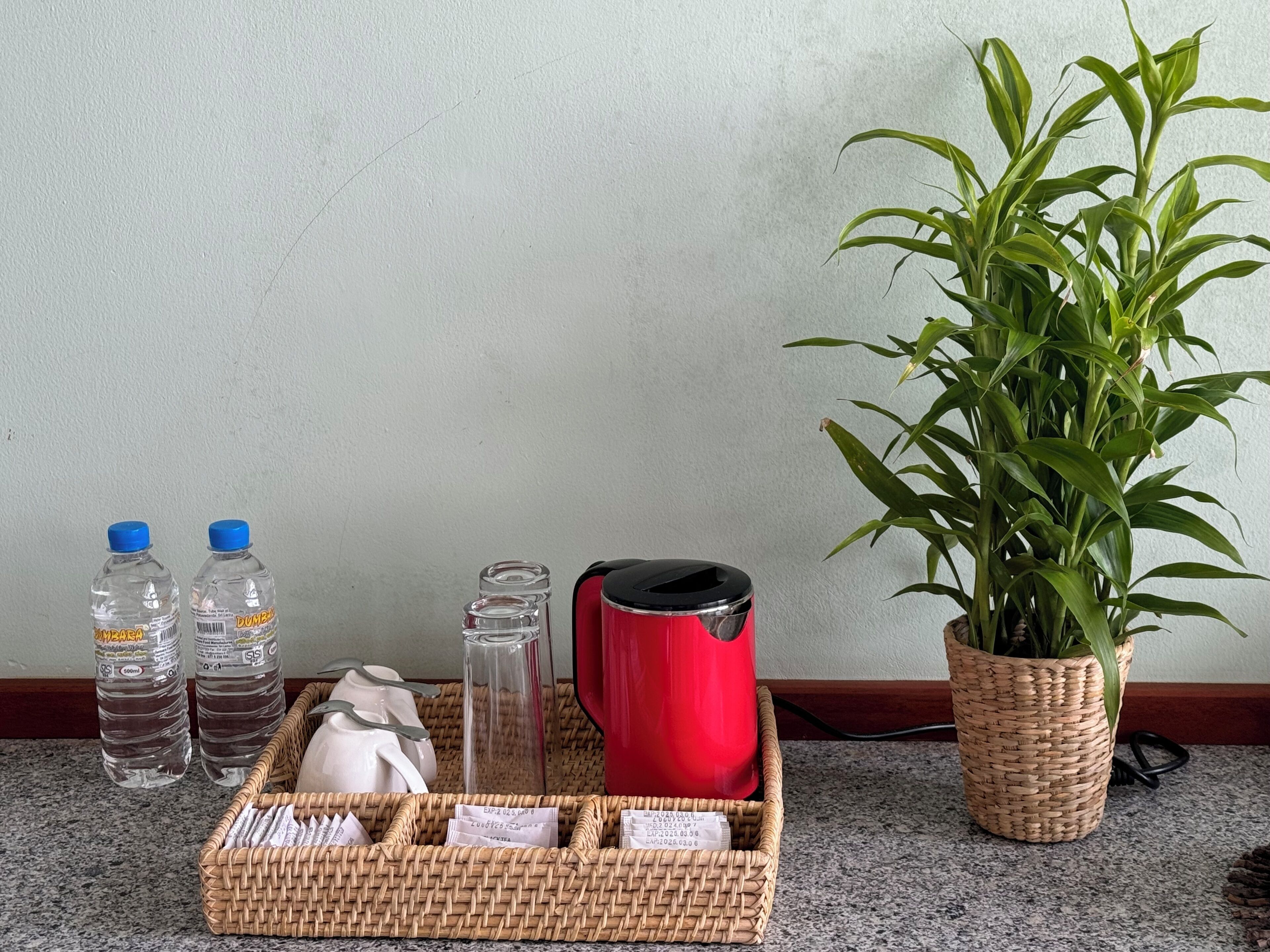 Uma bancada de cozinha com uma bandeja de vime contendo uma chaleira vermelha, duas garrafas de água, um jarro de vidro e uma pequena cesta com guardanapos e uma caixa de lenços. Uma planta em vaso também é visível.
