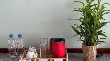 Uma bancada de cozinha com uma bandeja de vime contendo uma chaleira vermelha, duas garrafas de água, um jarro de vidro e uma pequena cesta com guardanapos e uma caixa de lenços. Uma planta em vaso também é visível.