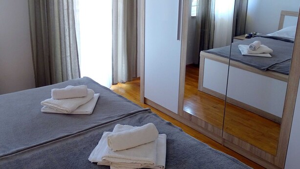 Quarto de hotel com duas camas, cada uma com um cobertor cinzento e toalhas brancas. Há um espelho na parede, e o chão é de madeira.